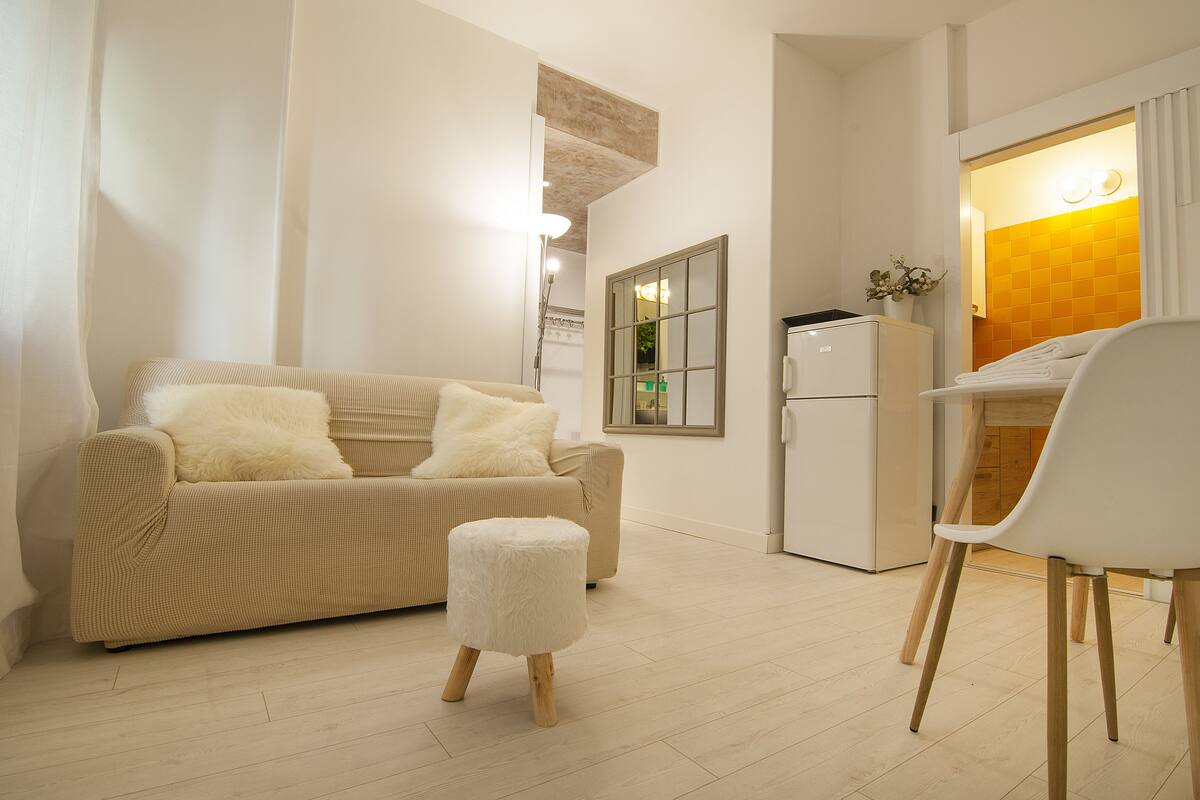 Uma sala de estar moderna com um sofá bege, um banquinho branco pequeno e uma área de jantar com uma cadeira e mesa brancas.