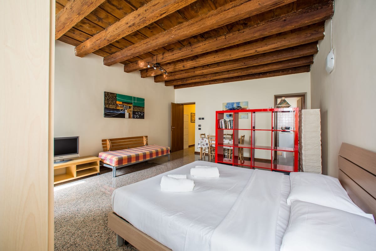 Quarto de hotel com teto de madeira, cama com roupa de cama branca, um armário vermelho, uma televisão e uma pequena mesa com uma cadeira.