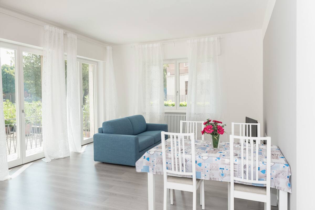 Um quarto claro com um sofá azul, uma mesa de jantar com cadeiras brancas e vista para uma varanda com cortinas brancas.