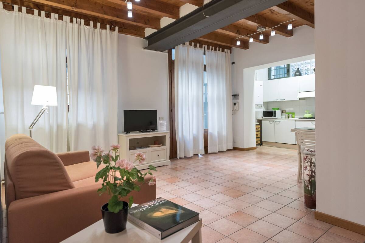 Uma sala de estar com um sofá, uma mesa de centro com um livro e flores, uma TV e uma cozinha ao fundo.