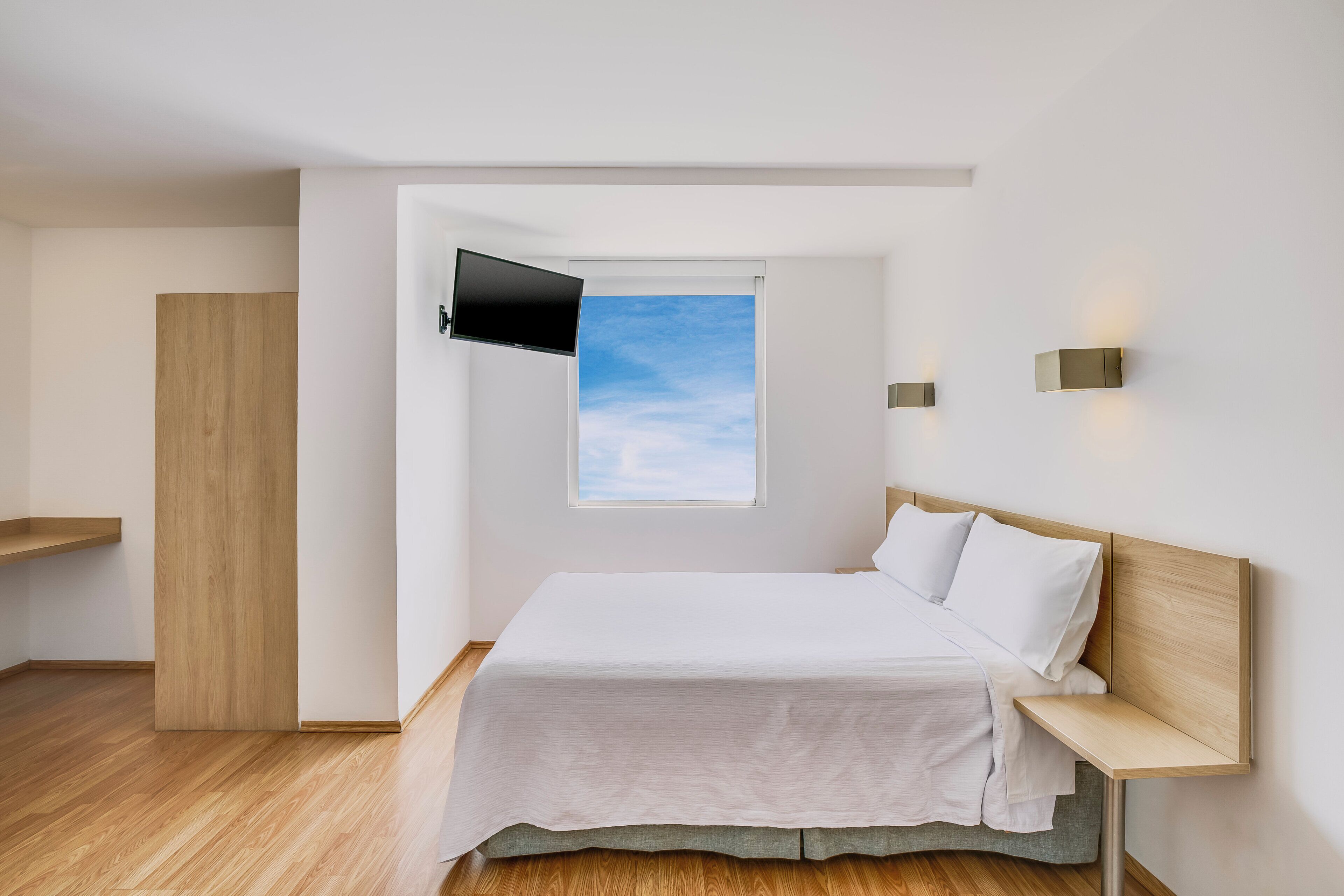 Quarto moderno com cama, TV fixada na parede e janela com vista para o céu.