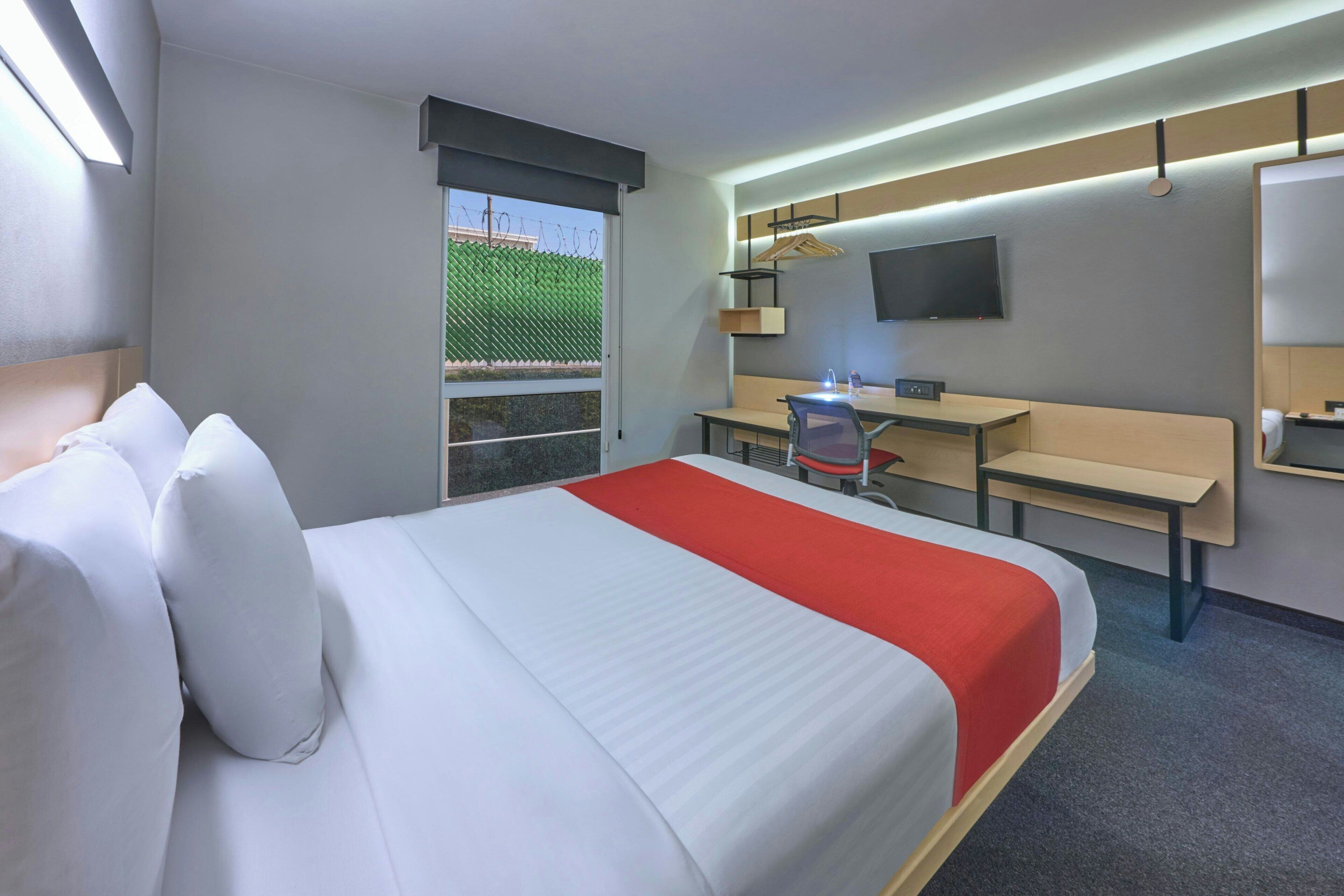 Quarto de hotel com cama, escrivaninha, cadeira, televisão e janela com vista para vegetação.