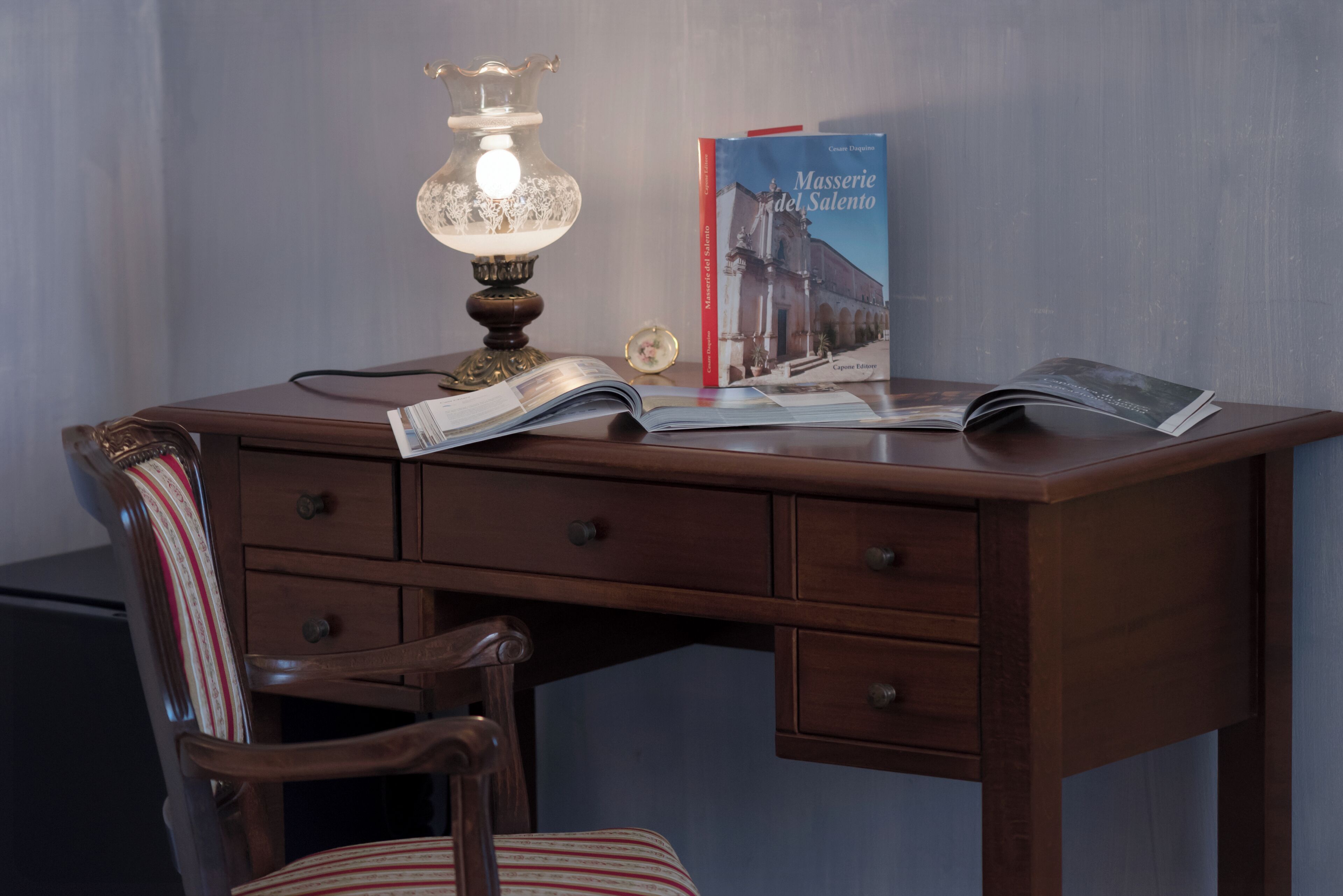 Uma mesa de madeira com uma lâmpada, um livro intitulado 'Masserie del Salento' e uma revista aberta.