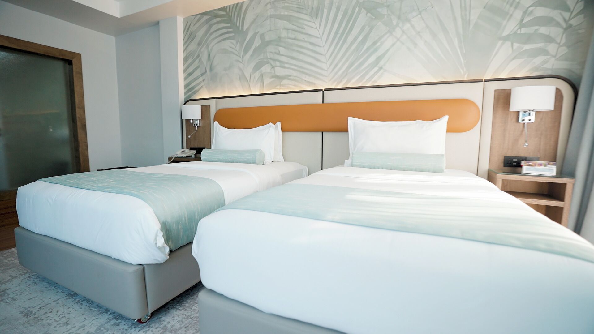 Quarto de hotel com duas camas, uma mesa de cabeceira, uma luminária e uma parede com padrão de folhas.