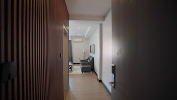 Um corredor com painel de madeira, área de estar com sofá e poltronas, e um ar condicionado fixado na parede.
