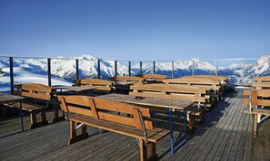 Um deck de madeira com mesas de piquenique e bancos, com vista para uma cadeia montanhosa coberta de neve.