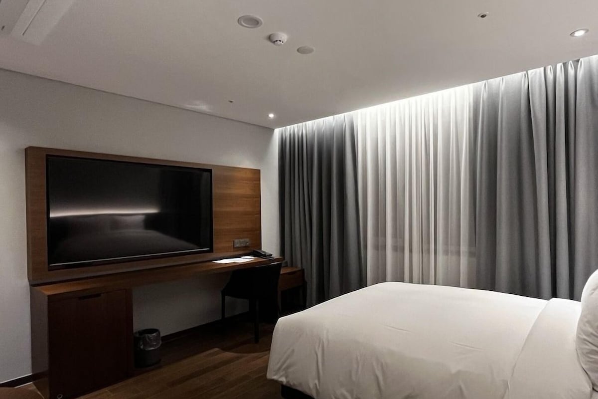 Quarto de hotel com cama, escrivaninha, televisão e uma grande janela com cortinas.