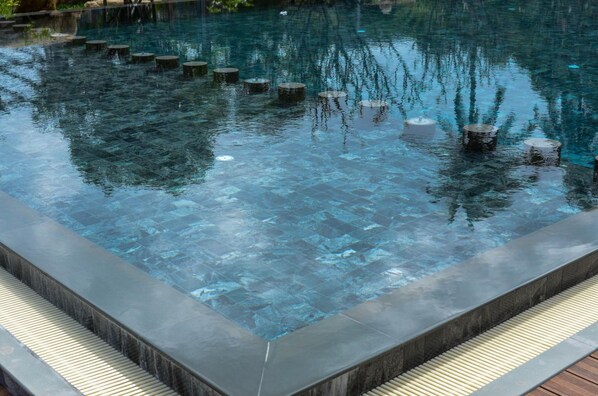 Piscina retangular com superfície de água azul translúcida, ladeada por uma borda de pedra e com pedras circulares para acesso.