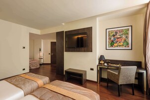 Quarto de hotel com cama, escrivaninha, cadeira, TV fixada na parede e um quadro emoldurado.