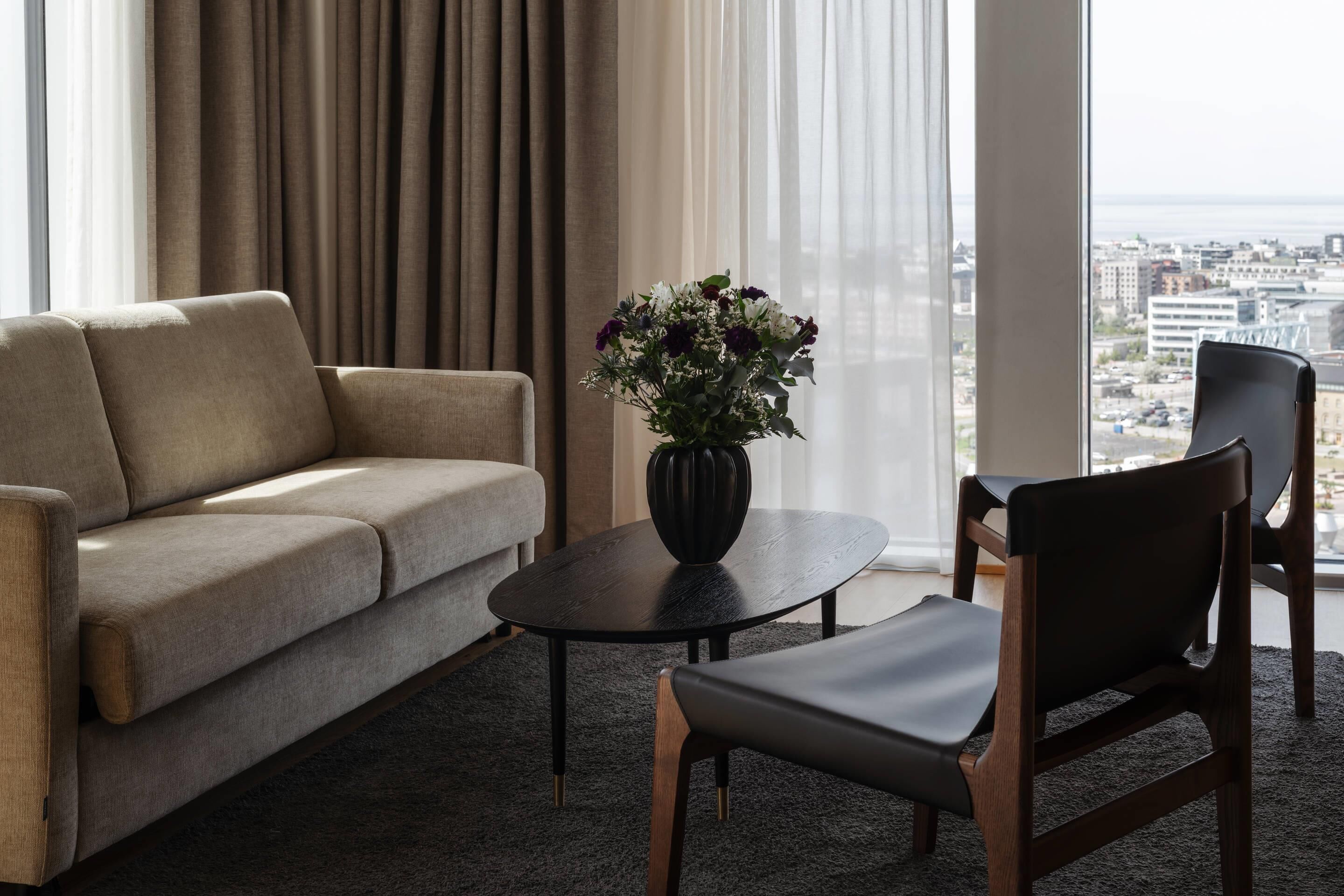 Uma sala de estar moderna com um sofá, uma mesa de centro e uma poltrona. Há um vaso com flores na mesa. O quarto tem uma janela grande com cortinas e vista para a paisagem urbana.