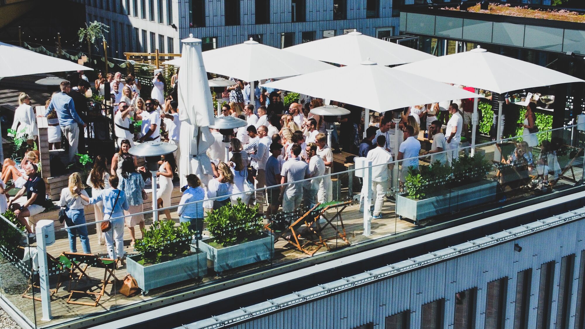 Um bar no terraço com mesas ao ar livre, guarda-sóis brancos e muitas pessoas.