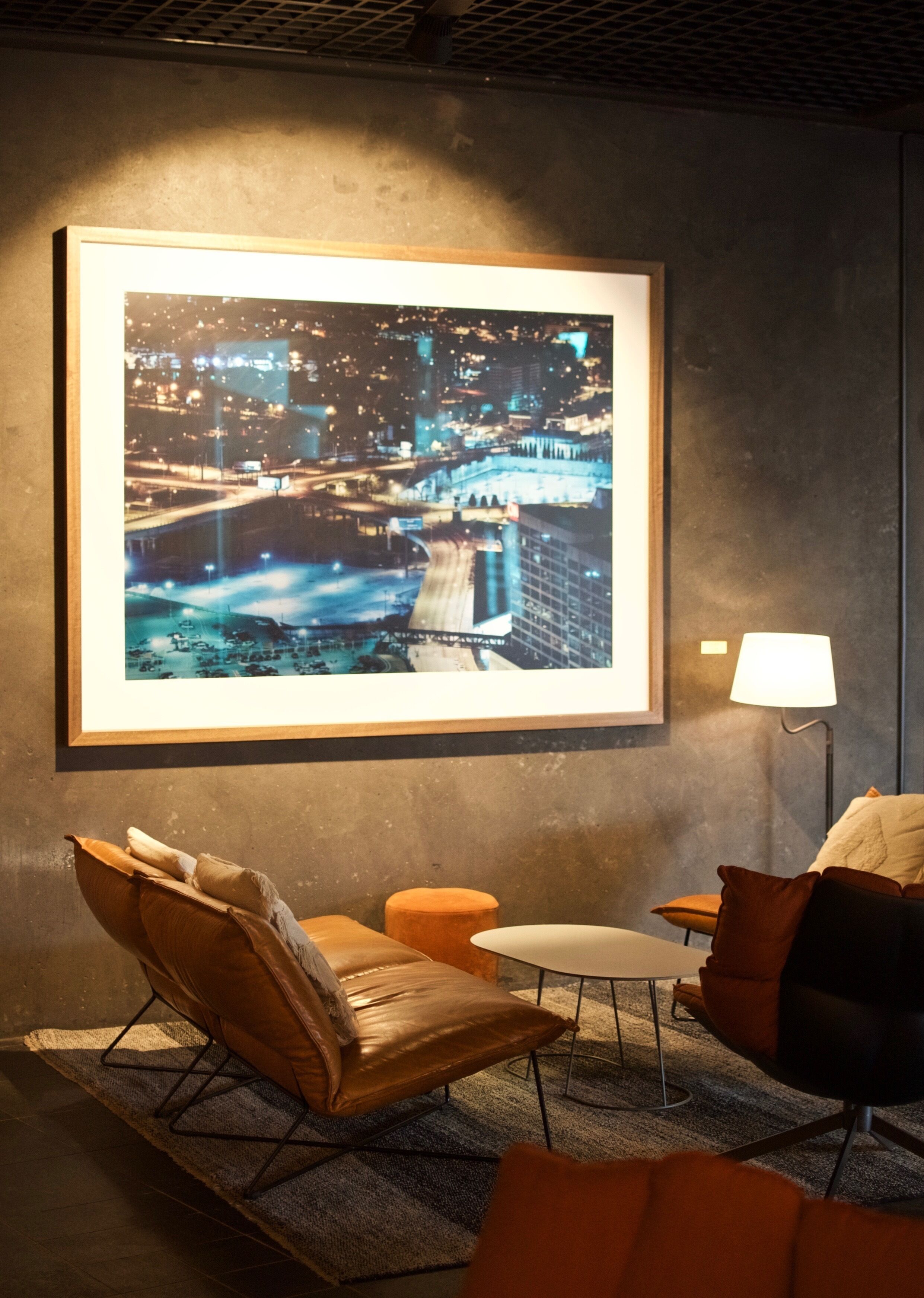 Uma sala de estar moderna com uma grande tela de pintura de paisagem urbana na parede, um sofá de couro, uma pequena mesa redonda e um abajur.