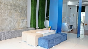 Um lobby moderno com um sofá bege, uma mesa de centro branca e um banco azul. Há painéis verticais verdes e uma cadeira esférica suspensa.