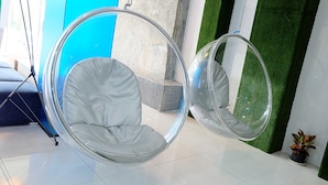 Duas cadeiras de balanço de plástico transparente com almofadas brancas em um quarto moderno, com esquema de cores azul e branco, uma parede de grama artificial verde e uma planta em vaso.