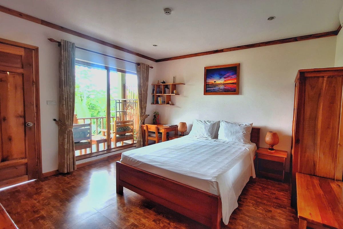 Quarto com cama grande, piso de madeira, um quadro na parede e uma varanda com vista para o verde.