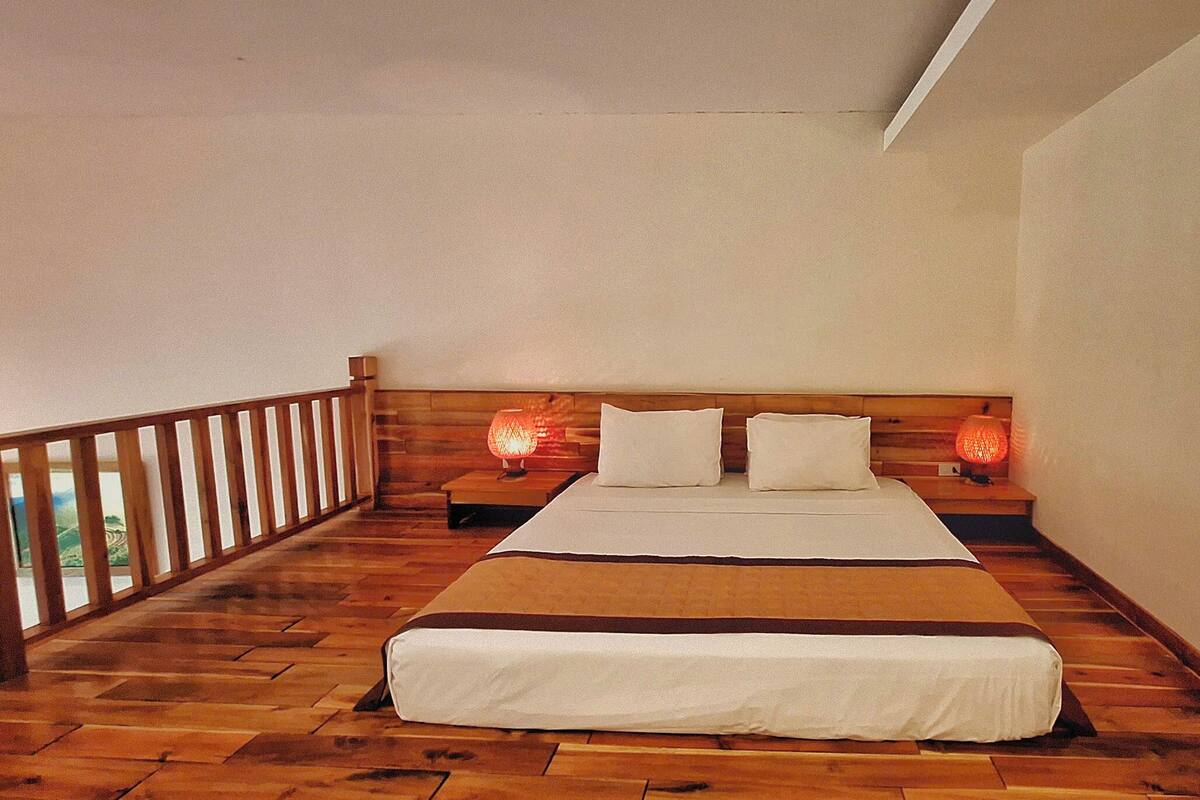 Uma cama grande com cabeceira de madeira, duas mesas de cabeceira e um corrimão de varanda.
