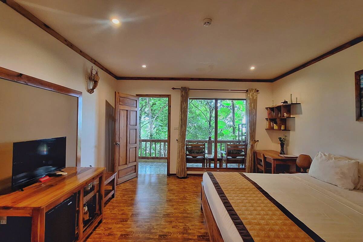 Quarto com piso de madeira, cama, televisão, escrivaninha e varanda com vista para o verde.