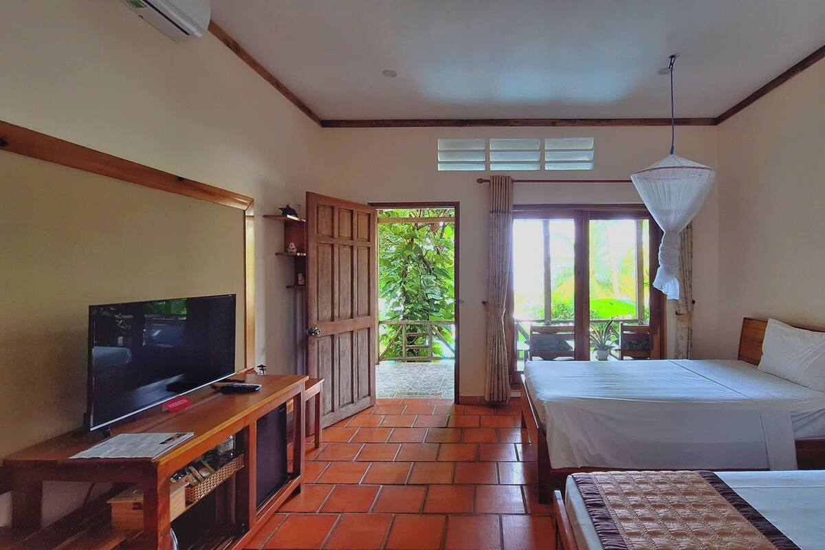 Quarto de hotel com cama, televisão, secretária e vista para o verde lá fora.