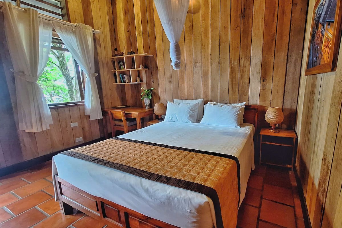 Quarto com painéis de madeira, uma cama, uma mesinha de cabeceira, uma estante com livros e uma planta, e uma janela com cortinas.