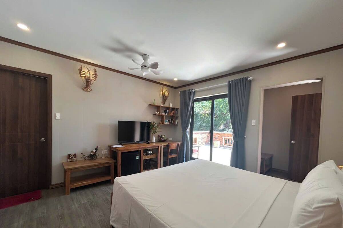 Quarto de hotel com uma cama grande, uma escrivaninha com televisão, um ventilador de teto e vista para o exterior por uma porta de correr.