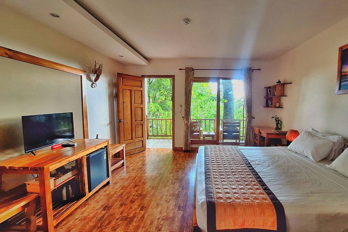 Quarto com cama, televisão, secretária de madeira e varanda com vista para o verde.