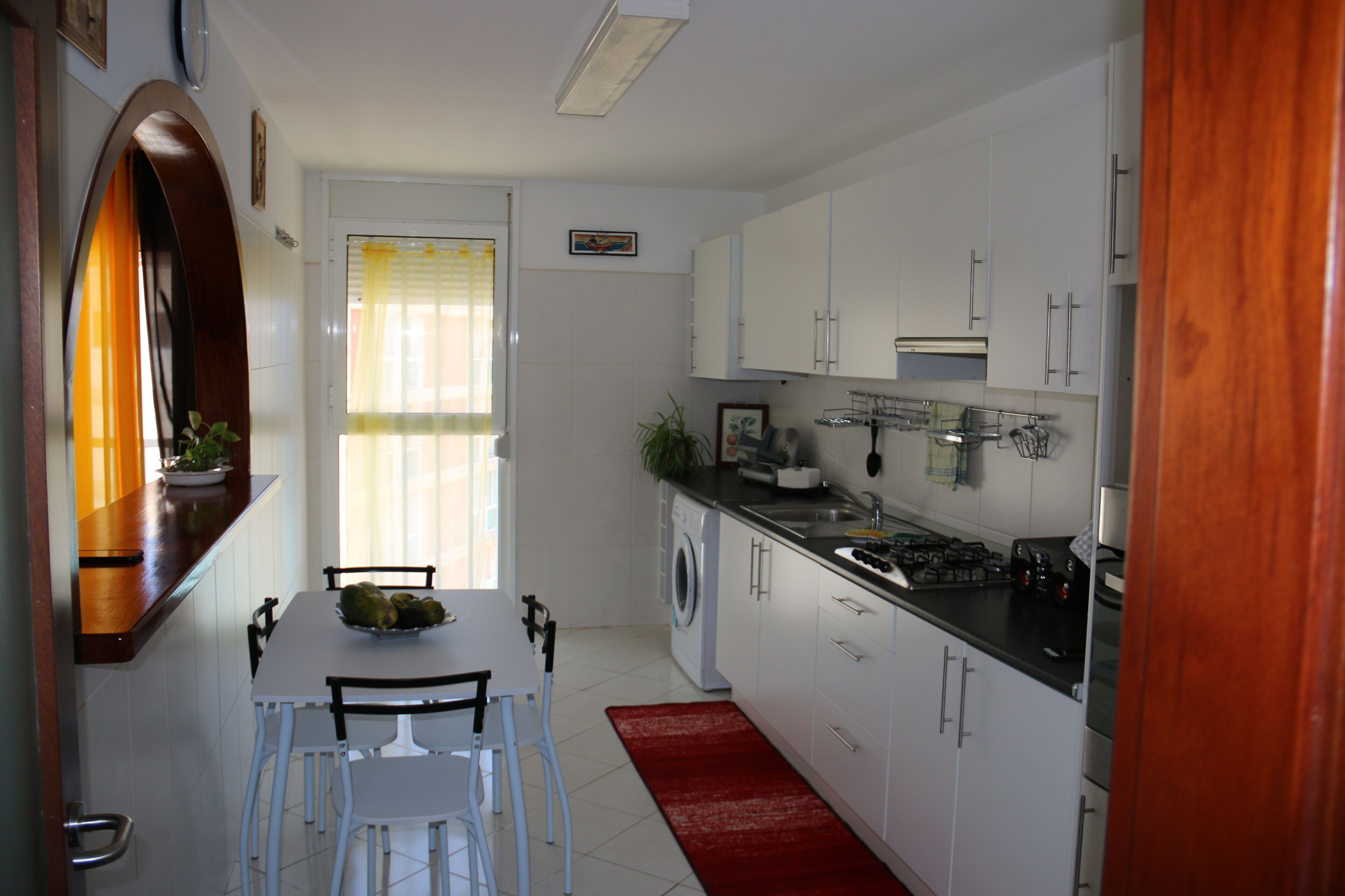 Cozinha compacta com armários brancos, máquina de lavar e área de jantar com mesa e cadeiras.