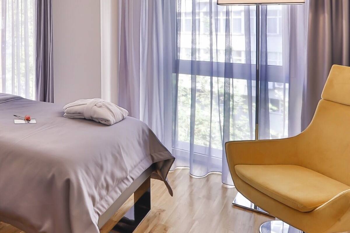 Quarto de hotel com cama, uma cadeira, abajur e janelas amplas com cortinas.