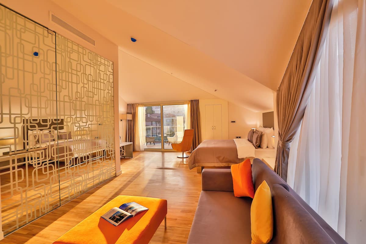 Quarto de hotel moderno com piso de madeira, sofá com almofadas laranja, cama, divisória de vidro e vista da cidade através de amplas janelas.