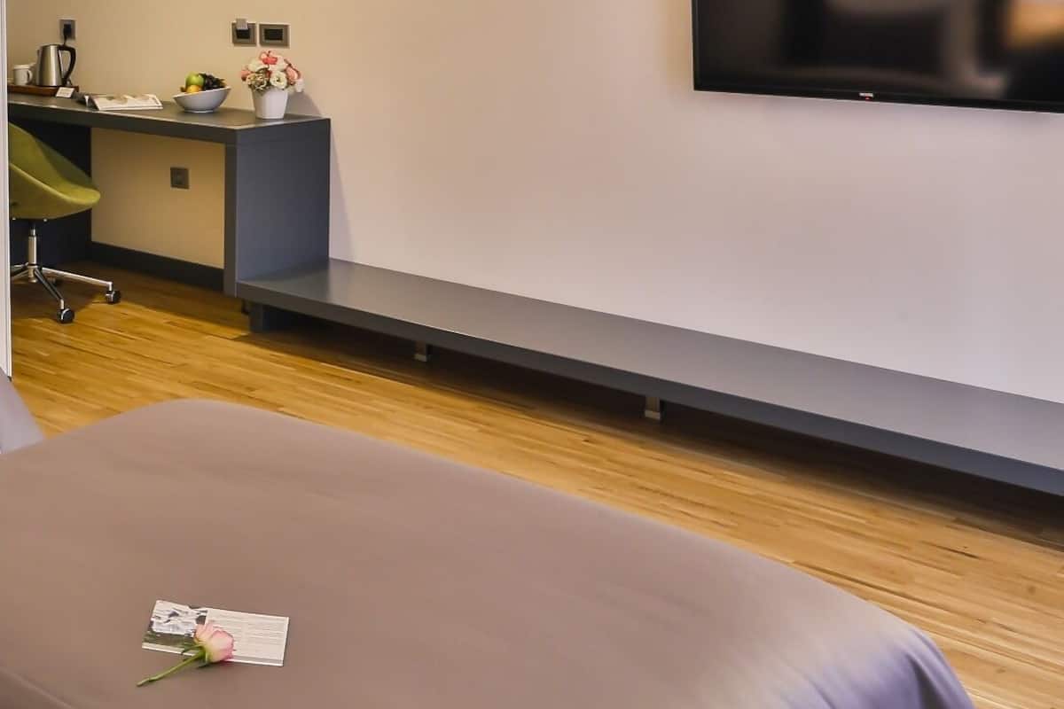 Quarto de hotel moderno com televisão de tela plana na parede, uma escrivaninha com chaleira e uma tigela de frutas, uma cadeira verde e uma cama com uma rosa.