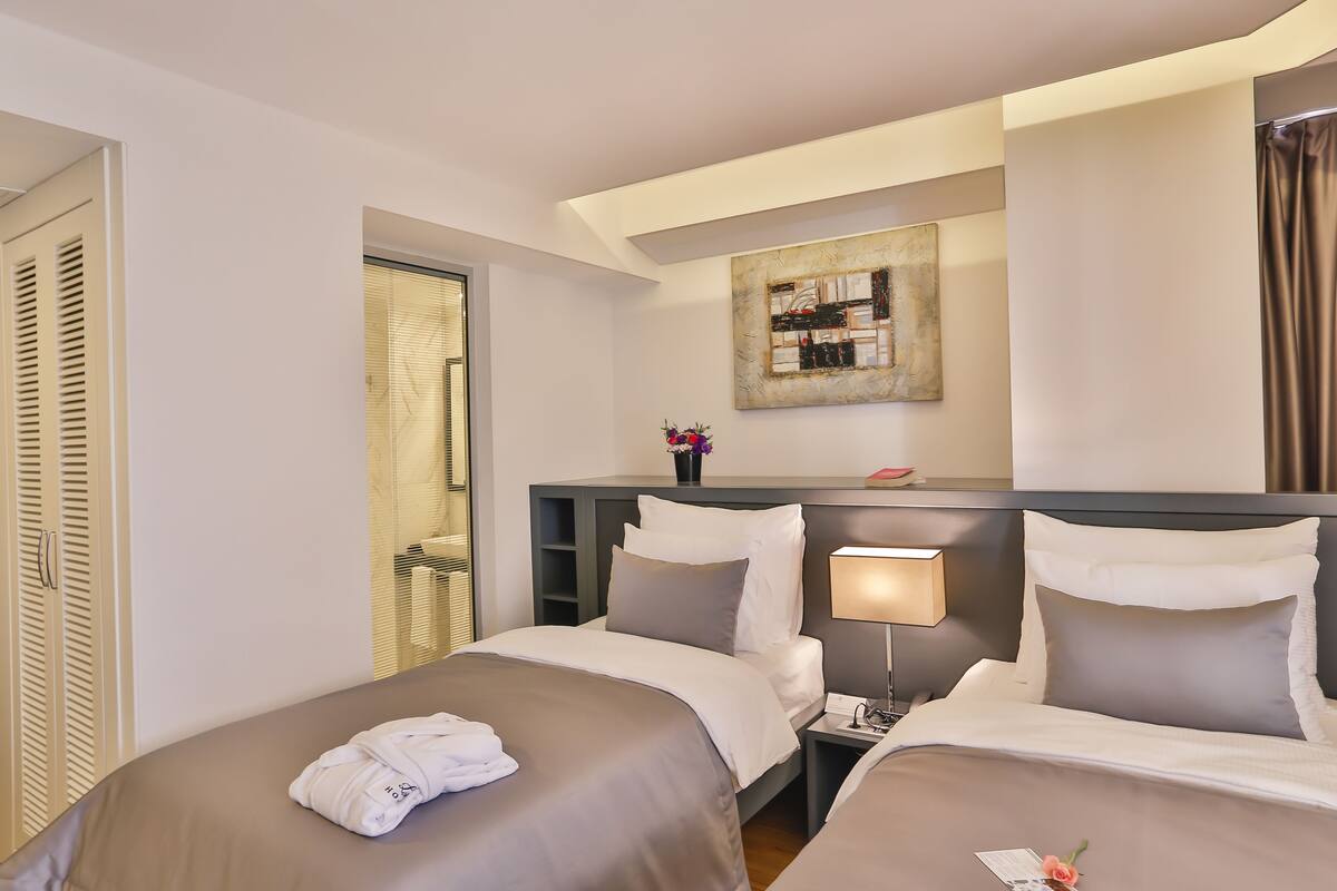 Quarto de hotel com duas camas, uma mesa de cabeceira com abajur, um espelho e uma obra de arte na parede.