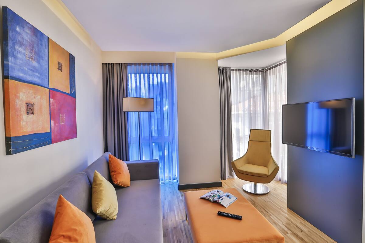 Quarto de hotel moderno com sofá, puf e poltrona, televisão de tela plana e uma grande janela com cortinas.