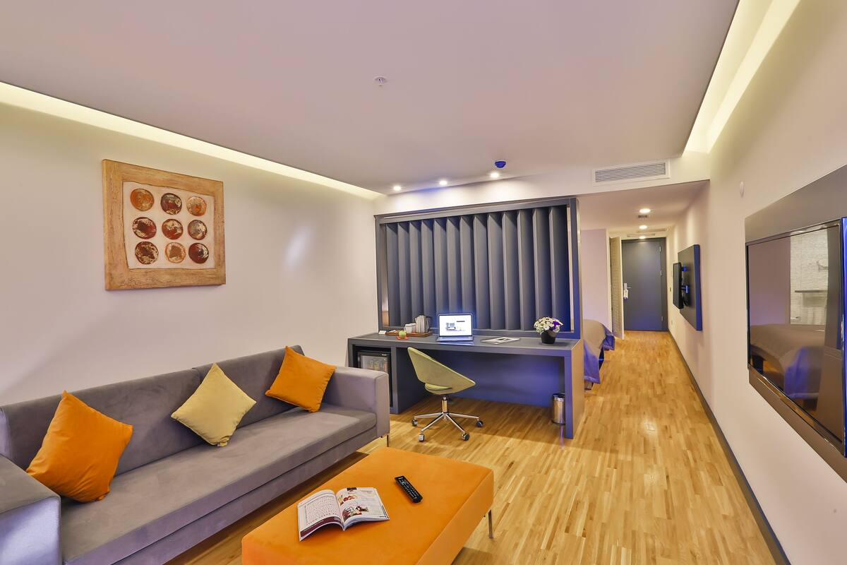 Quarto de hotel moderno com sofá cinza, almofadas laranja, piso de madeira, escrivaninha com laptop, televisão e uma obra de arte emoldurada na parede.