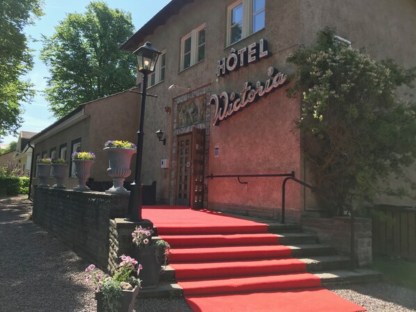 Um hotel com entrada carpetada de vermelho, uma placa indicando "HOTEL Victoria" e grandes vasos de flores nos degraus.