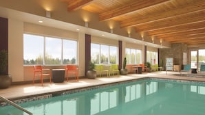 Piscina com borda azulejada, rodeada por cadeiras e mesas, e grandes janelas com vista para o exterior.