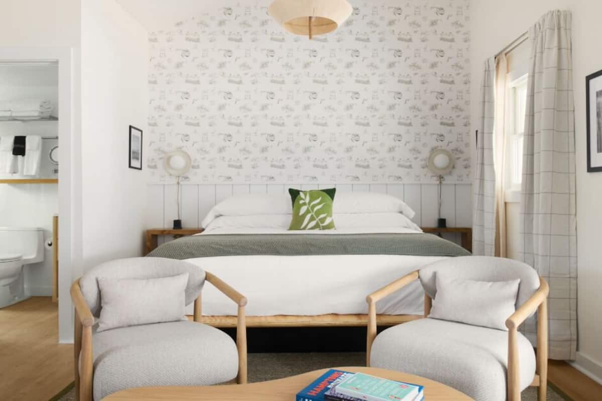 Quarto com cama, duas cadeiras, mesa, abajur, janela com cortinas e parede com papel de parede estampado.