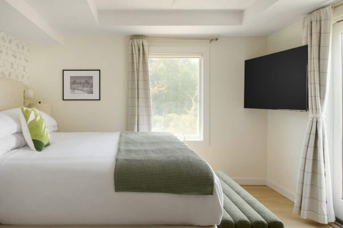 Quarto com cama, luminária de cabeceira, janela com cortinas, televisão na parede e quadro emoldurado.
