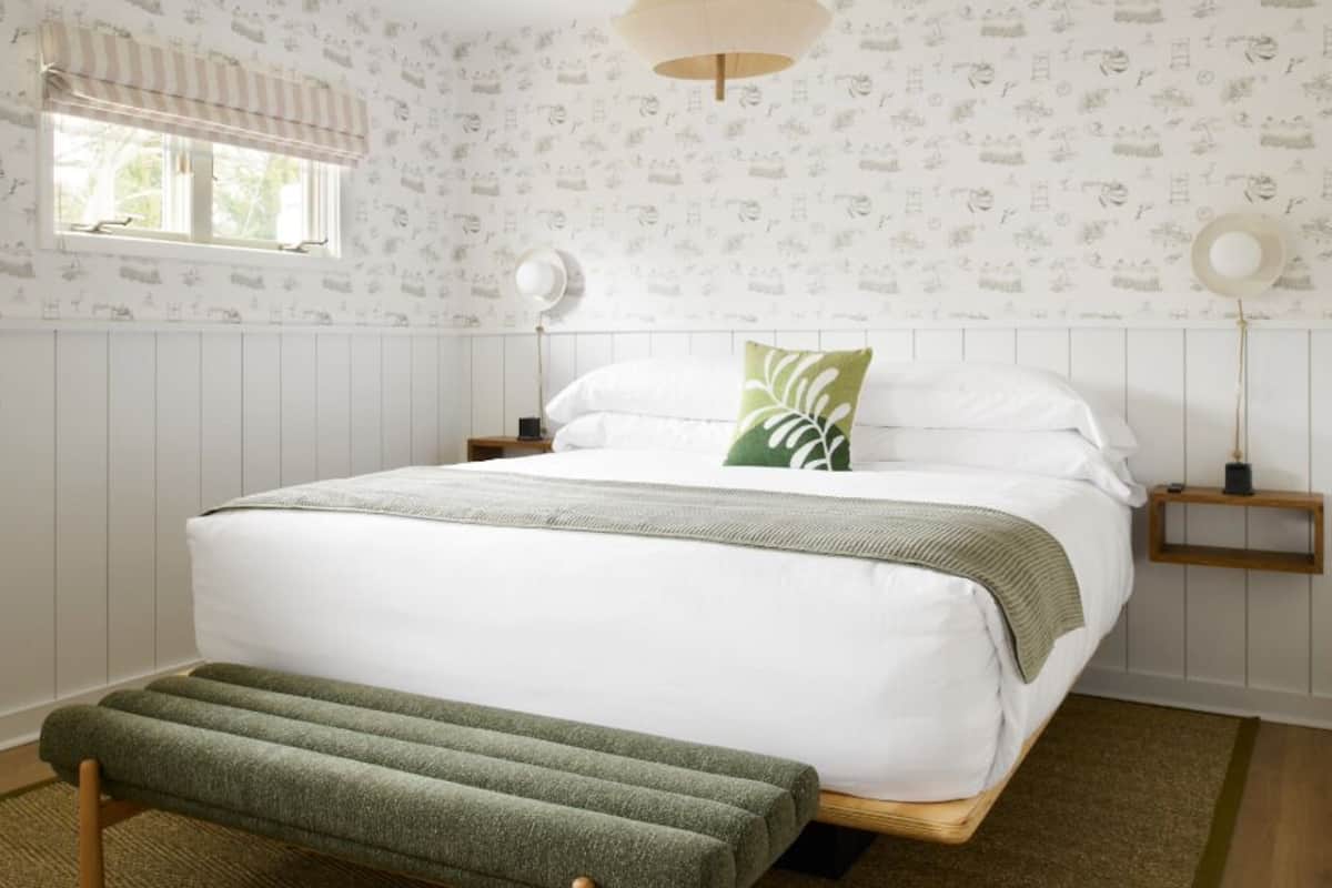 Quarto com cama grande, um banco, janela com cortina e parede com padrão.