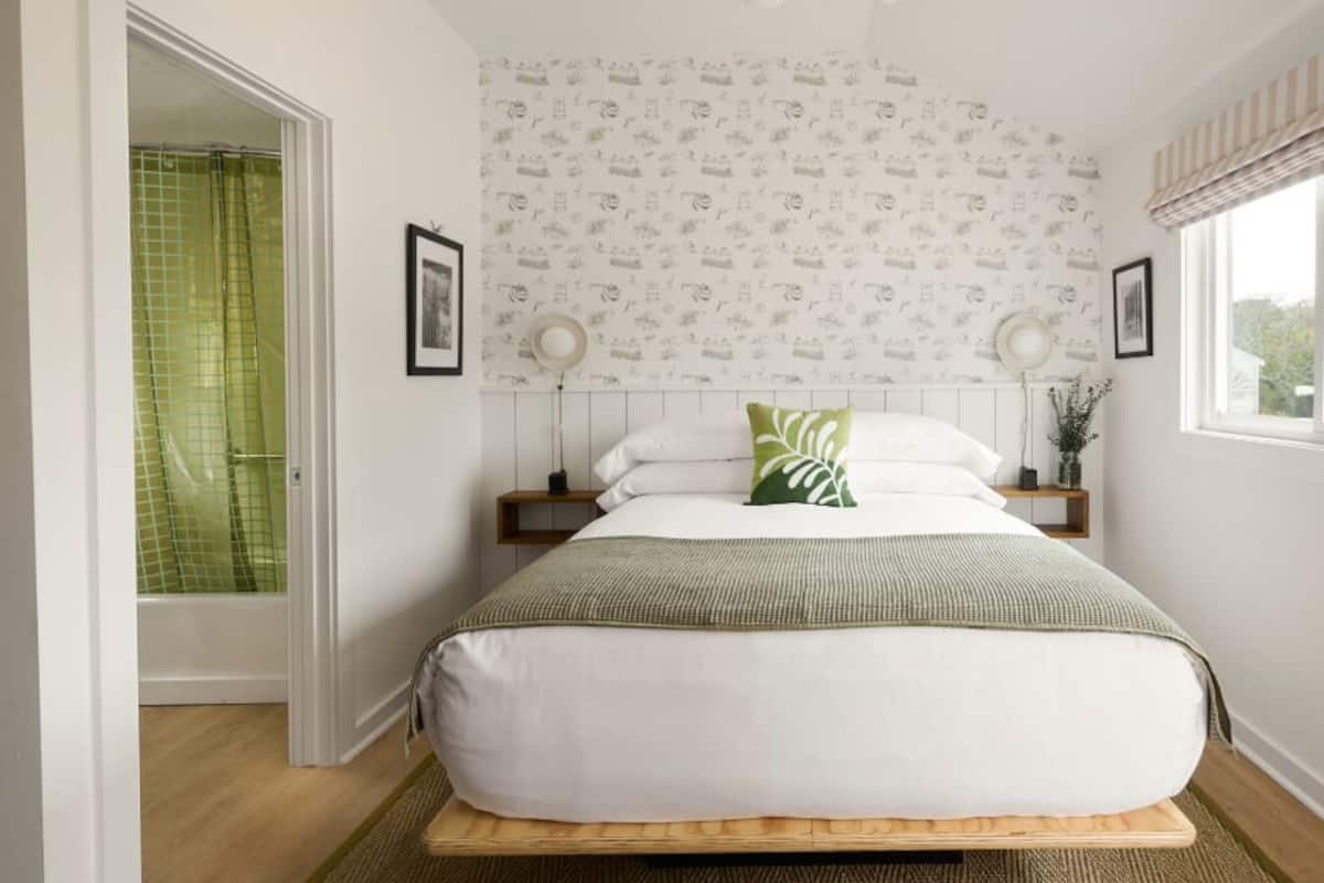 Quarto com cama centralizada em uma plataforma, duas mesas de cabeceira com luminárias, uma janela com cortinas e papel de parede estampado.