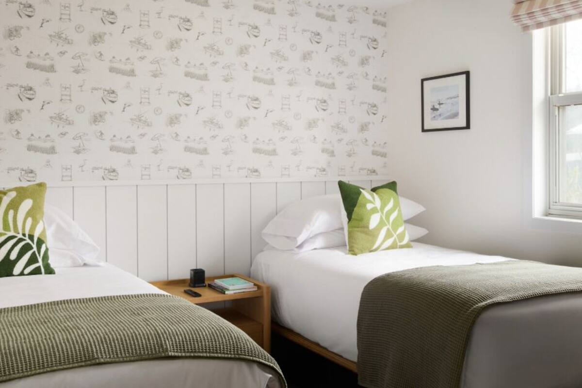 Quarto de hotel com duas camas, uma mesa de cabeceira com abajur, um quadro na parede e uma janela com cortinas.