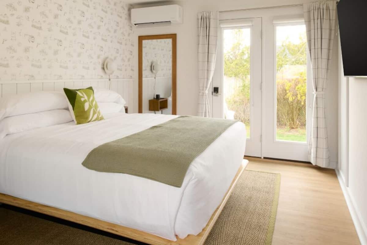 Quarto com cama, mesa de cabeceira, espelho, janela com cortinas e porta para o exterior.