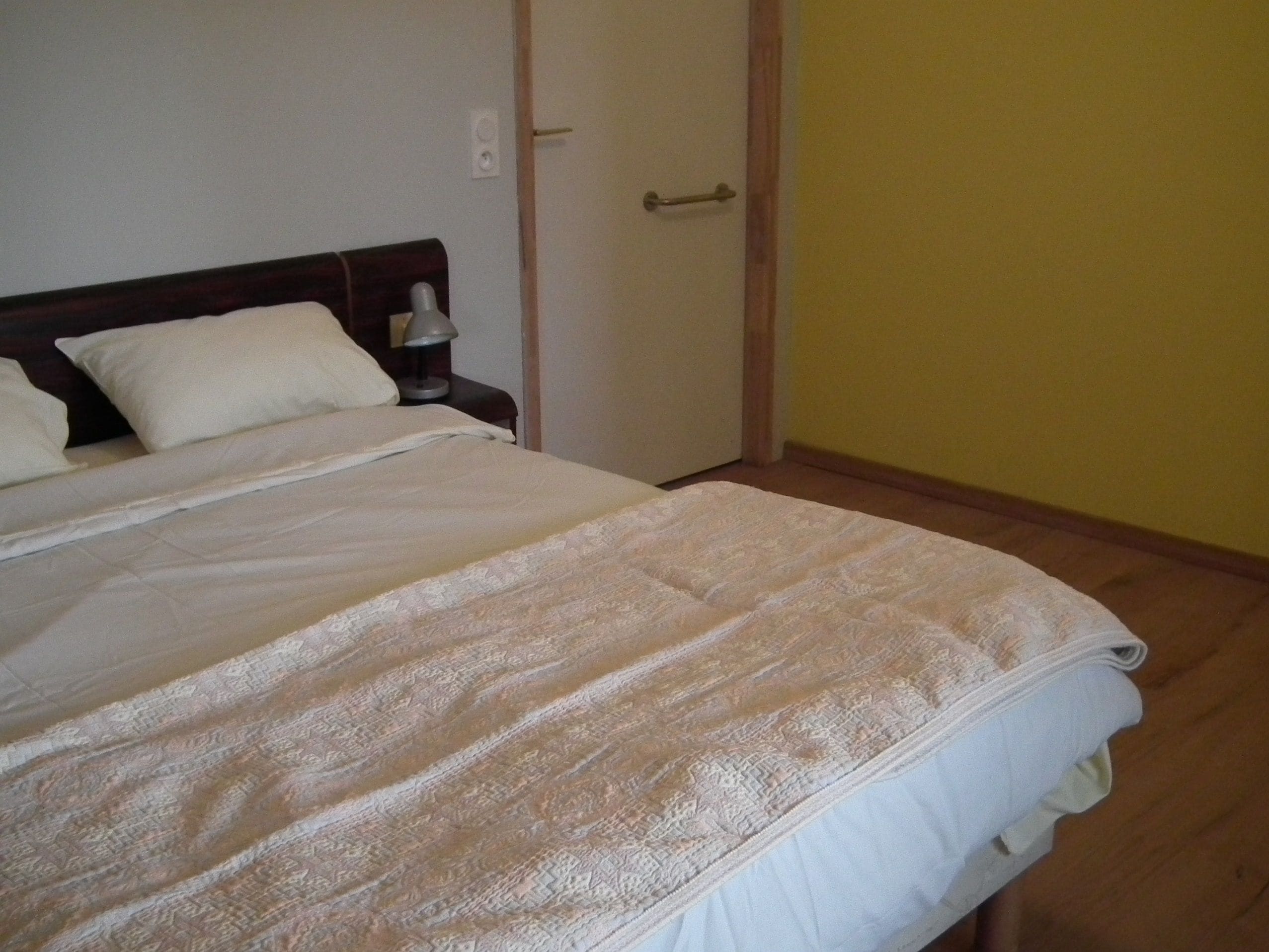 Uma cama com colchão branco e manta estampada, uma mesa de cabeceira com abajur e uma porta com puxador.