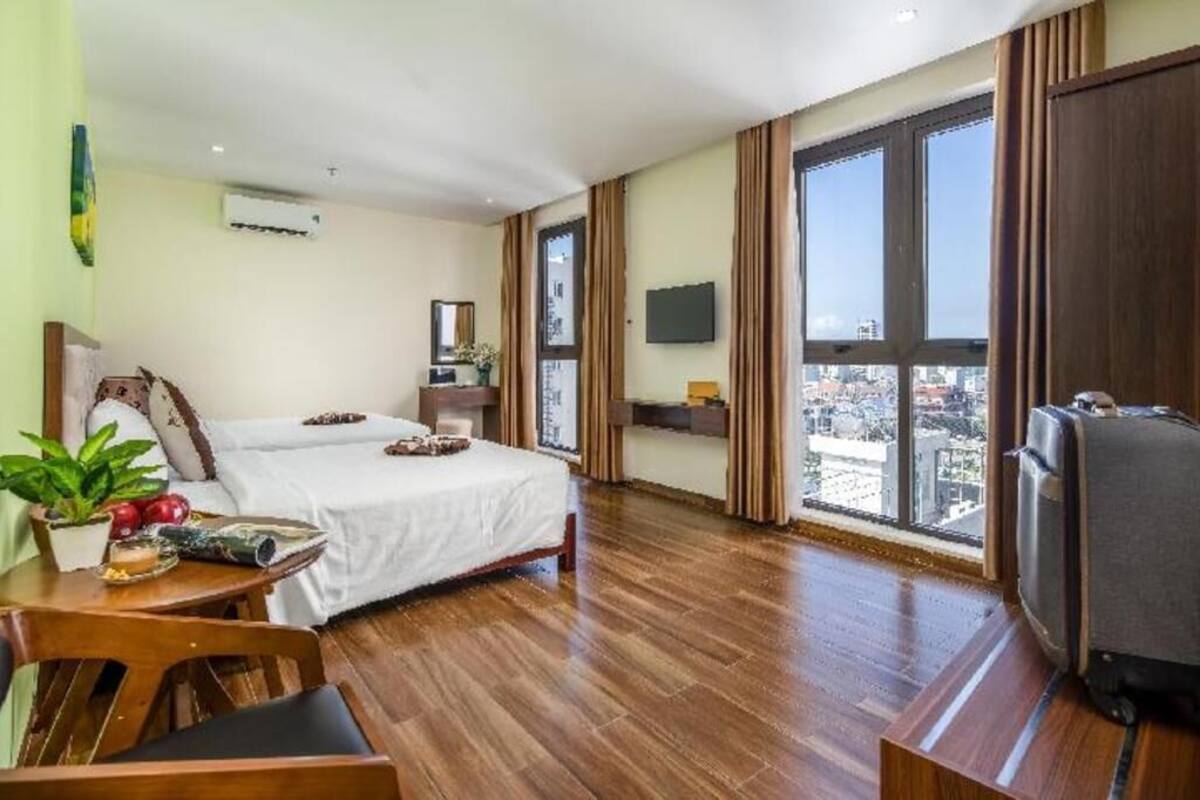 Quarto de hotel com cama, escrivaninha com cadeira, televisão, janela grande com cortinas e vista para a cidade.
