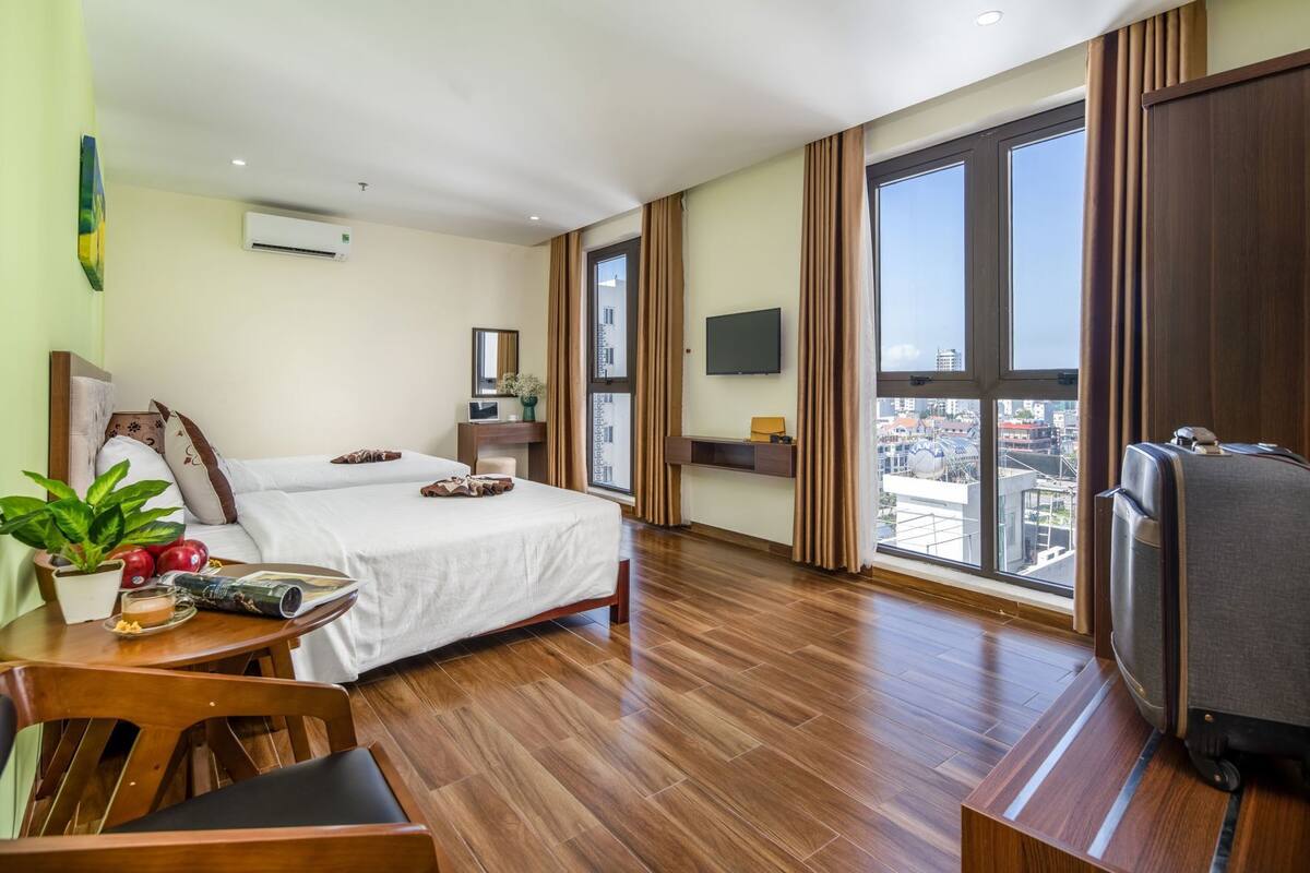 Quarto de hotel com cama, escrivaninha com planta, cadeira, mala, televisão e janela ampla com vista para a cidade.
