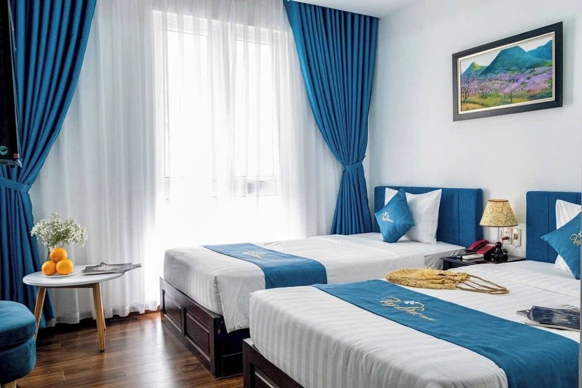 Quarto de hotel com duas camas, uma mesa pequena com um vaso de flores e um quadro na parede.
