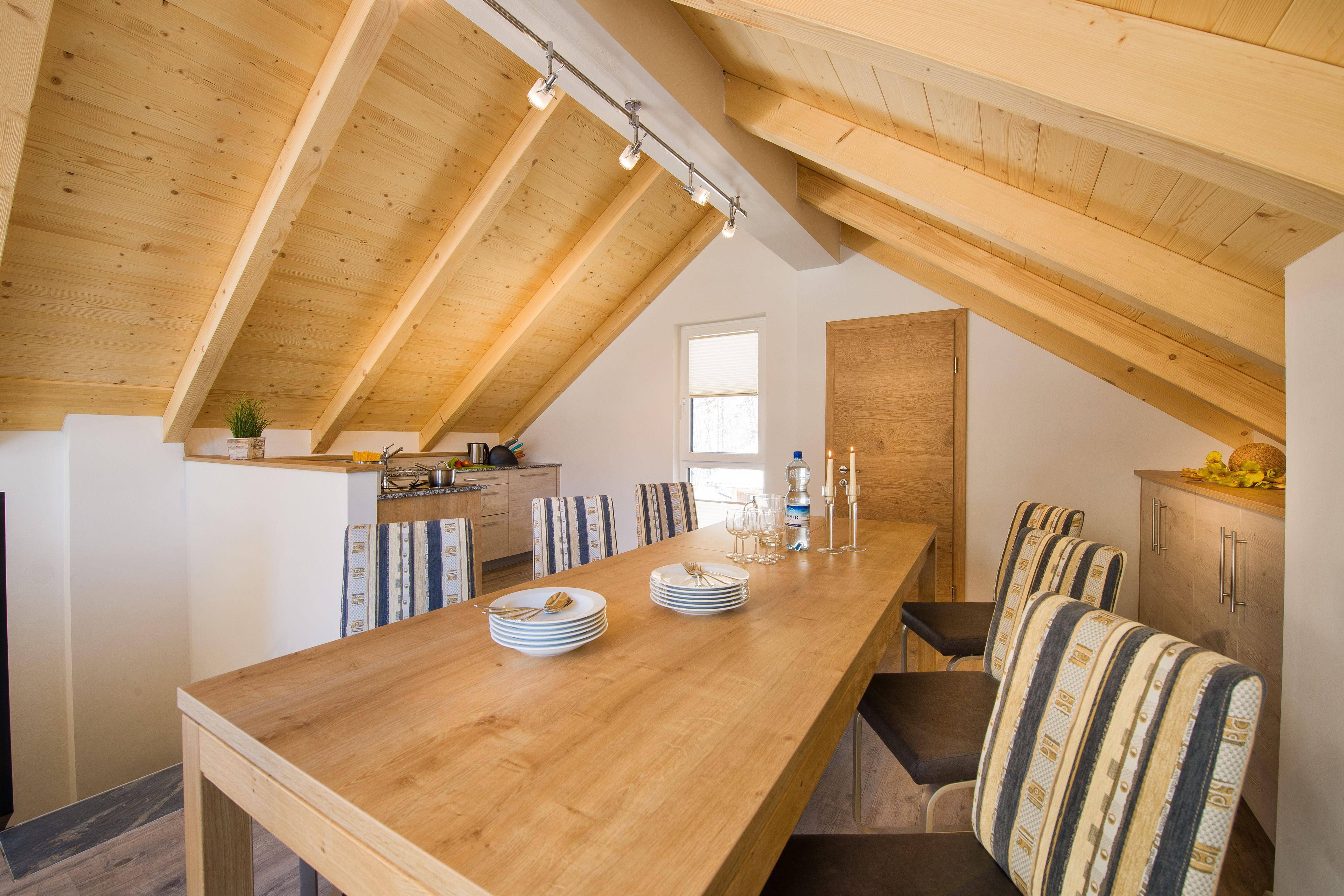 Uma área de refeições com uma mesa de madeira, cadeiras listradas e uma cozinha ao fundo.