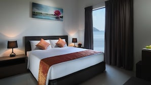 Quarto de hotel com uma cama grande, duas mesas de cabeceira com luminárias, um quadro na parede, vista para o mar pela janela e uma cesta de frutas numa mesinha lateral.