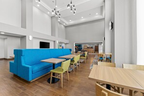 Interior moderno com sofá azul, mesas e cadeiras de madeira, e uma cozinha ao fundo.