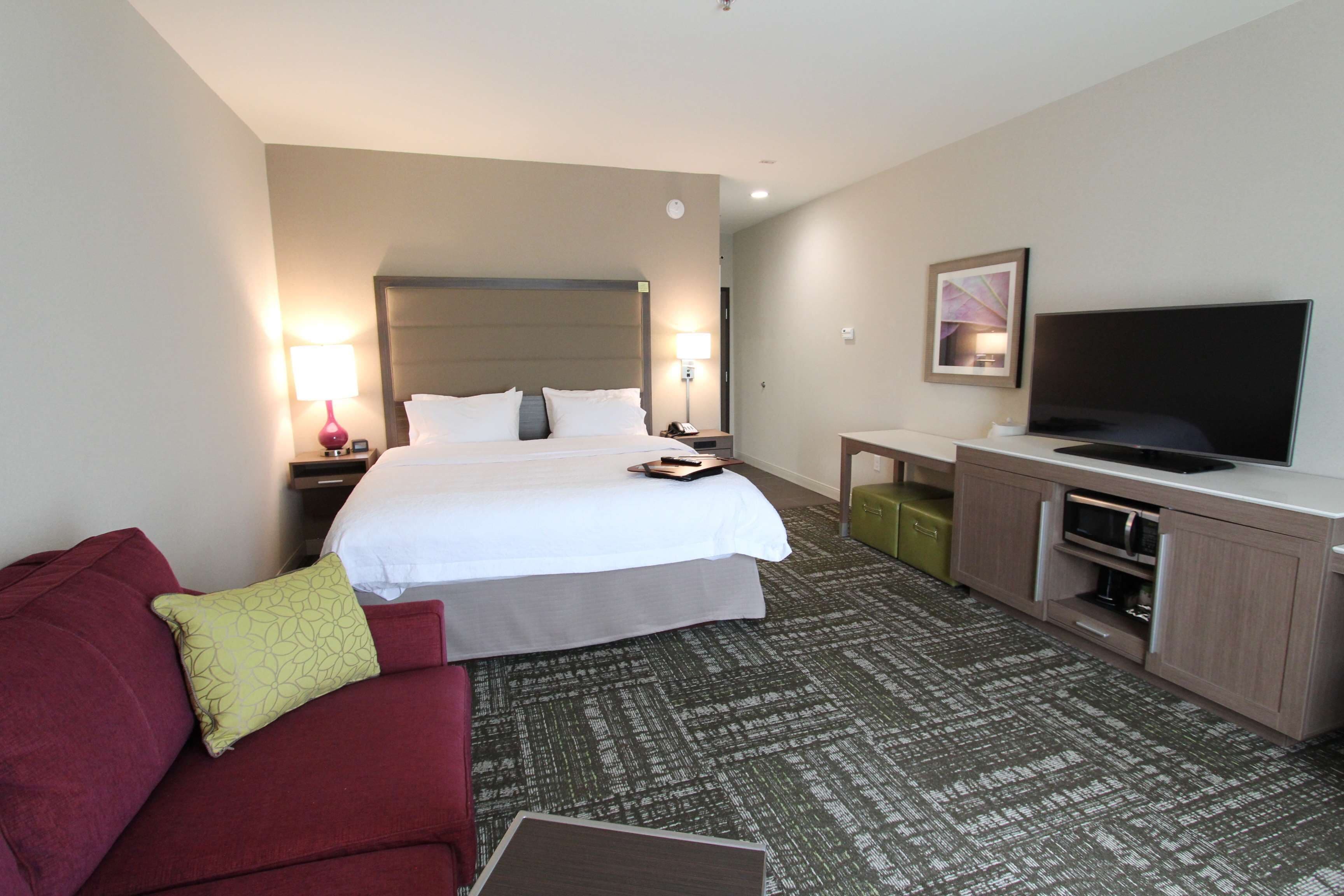 Quarto de hotel com cama, televisão, escrivaninha, sofá e piso coberto por carpete.
