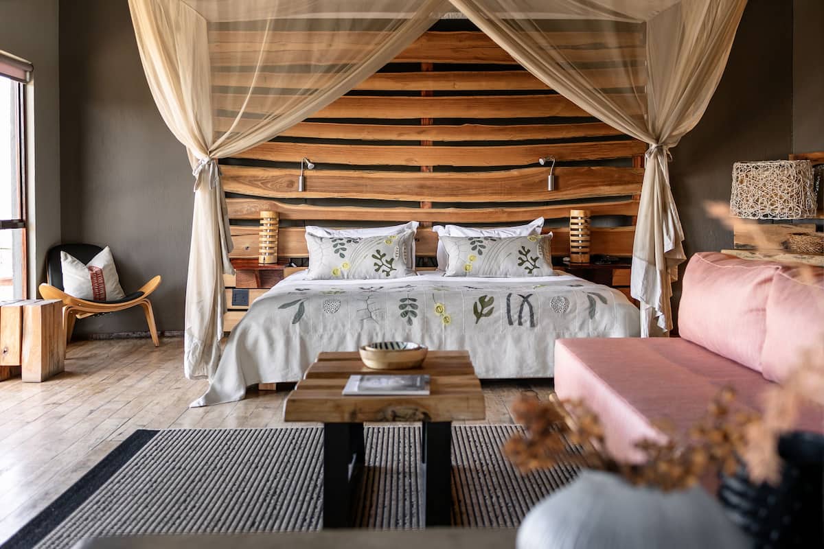 Quarto com cabeceira de madeira, cama com dossel, mesa de cabeceira com uma tigela, sofá rosa e tapete de fibras.