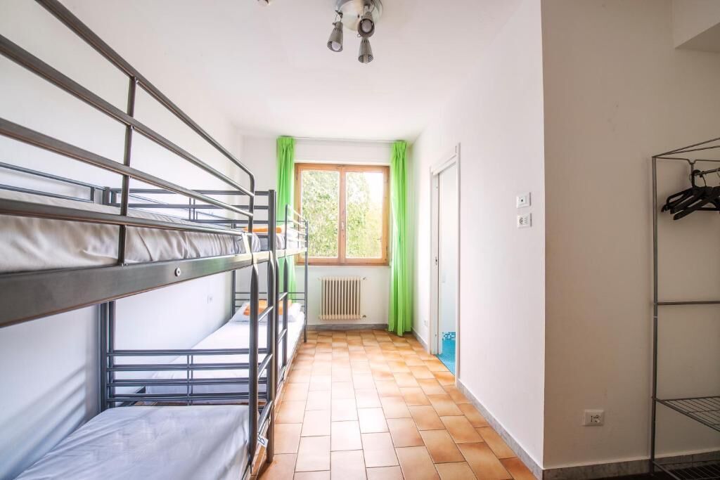 Um quarto de dormitório com beliches, um radiador, uma janela com cortinas e uma estante de metal com roupas penduradas.